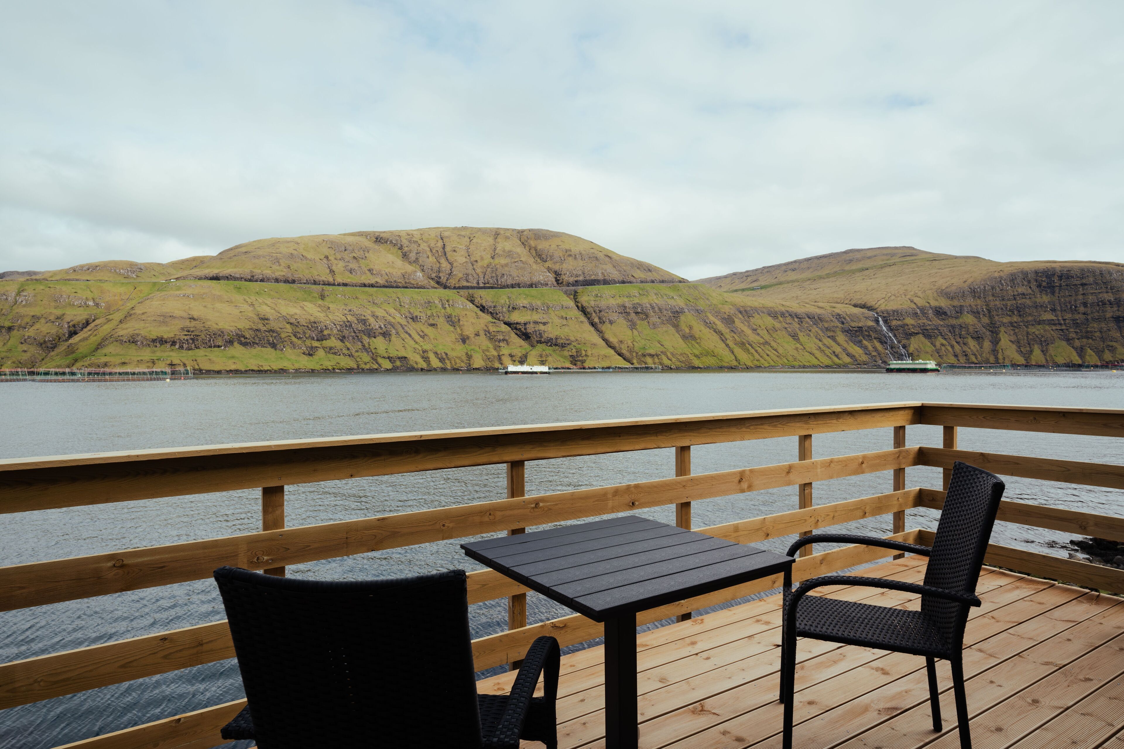 Um deck de madeira com uma mesa e cadeiras, dando para um lago com colinas ao fundo.