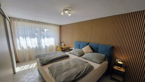 Quarto moderno com uma cama grande, parede de ripas de madeira, escrivaninha com cadeira e uma janela ampla com cortinas leves.