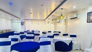 Uma sala de banquetes com mesas e cadeiras redondas, toalhas de mesa brancas e azuis, e luminárias pendentes.