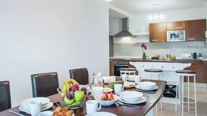 Uma área de jantar moderna com uma mesa de madeira posta para o café da manhã, incluindo frutas, doces e bebidas. Uma cozinha com eletrodomésticos de aço inoxidável e armários de madeira é visível ao fundo.