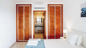 Quarto de hotel com cama, mesa de cabeceira, luminária e banheiro visível por uma porta aberta.