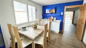 Uma área de refeições com mesa e cadeiras de madeira, televisão fixada numa parede azul e cozinha ao fundo.