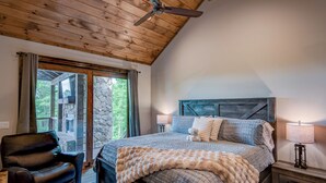 Quarto com teto de madeira, cama com cabeceira azul, mesa de cabeceira com abajur e vista para o exterior através de uma porta de vidro.