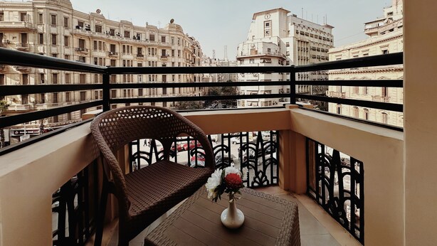 Varanda com cadeira de vime e um vaso de flores, com vista para um horizonte urbano repleto de edifícios históricos.