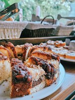 Uma tábua de pão fatiado com diversos acompanhamentos, um par de pinças, e uma bandeja de doces ao fundo.