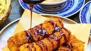 Um prato com petiscos empanados e fritos, regados com molho escuro, acompanhado de uma tigela de molho e uma cesta de pão fatiado.