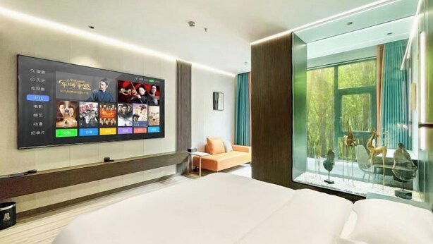 Quarto de hotel com uma televisão de tela grande fixada na parede, uma cama, um sofá e uma janela ampla com vista para o exterior.