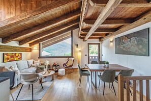 Um loft espaçoso com teto alto, vigas de madeira, uma grande mesa de jantar com cadeiras, um sofá e uma poltrona com um vaso de flores.