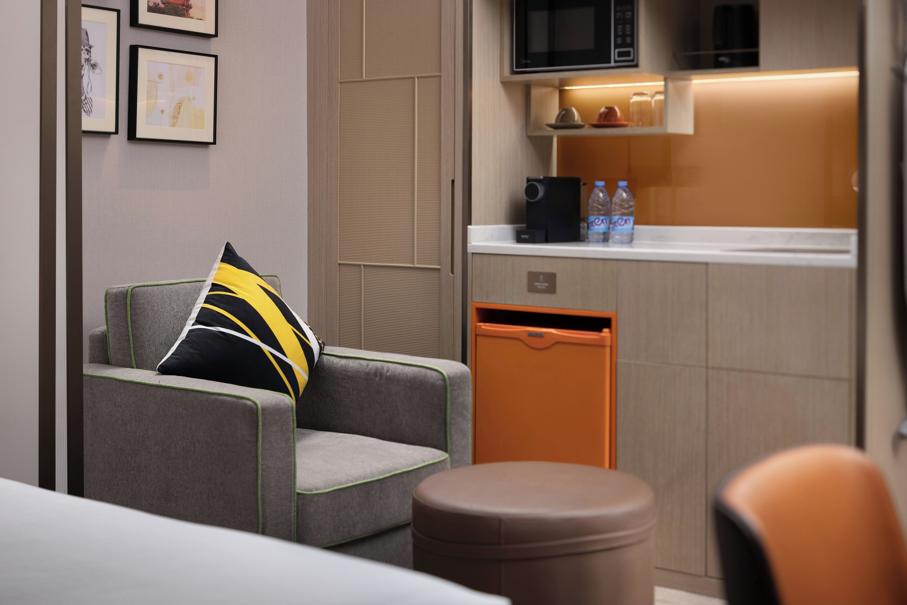 Quarto moderno com sofá cinzento, um banquinho redondo pequeno, cozinha compacta com micro-ondas e um cesto de lixo.