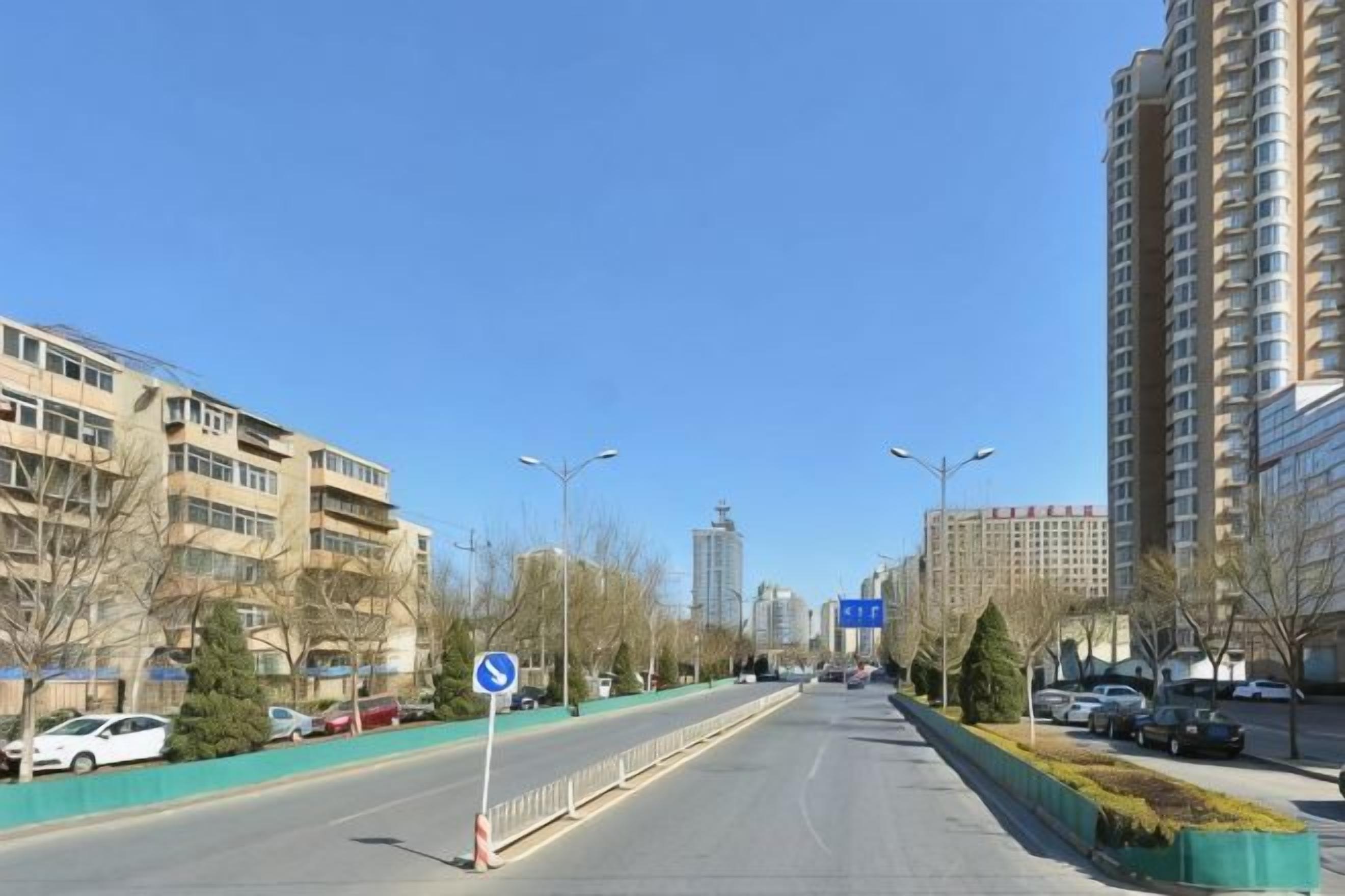 Uma rua urbana ampla com edifícios altos, céu claro e uma placa azul indicativa.