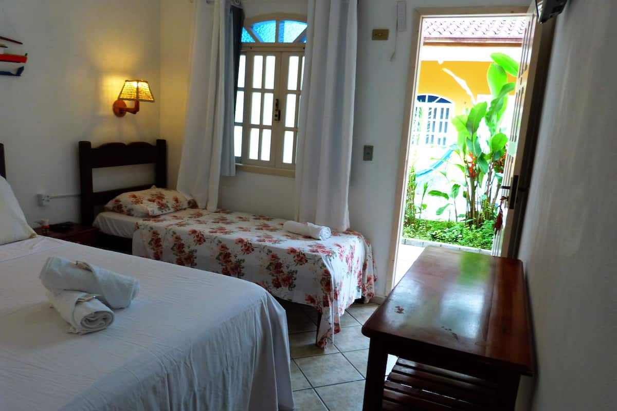 Quarto de hotel com duas camas, uma mesinha de cabeceira de madeira, uma mesa, uma janela com cortinas e vista para o verde lá fora.
