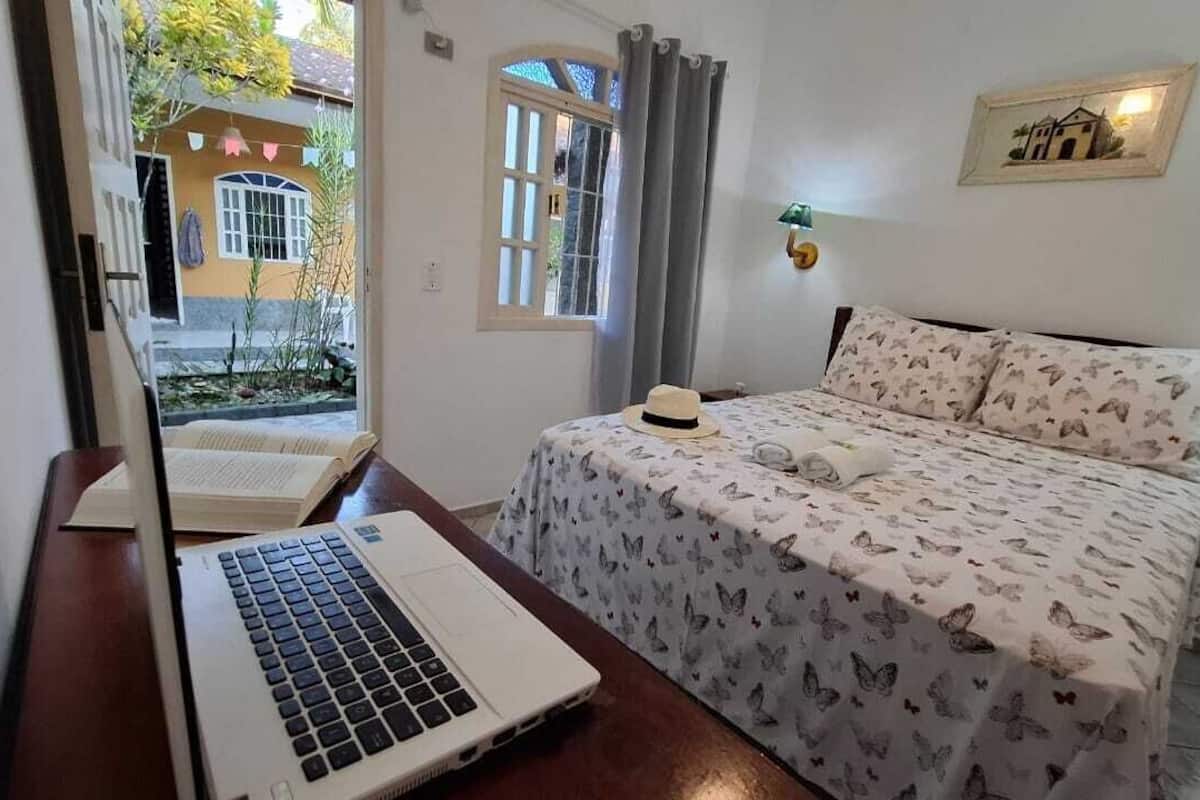Um quarto com uma cama, um laptop sobre uma escrivaninha, uma janela e uma porta que dá para o exterior.