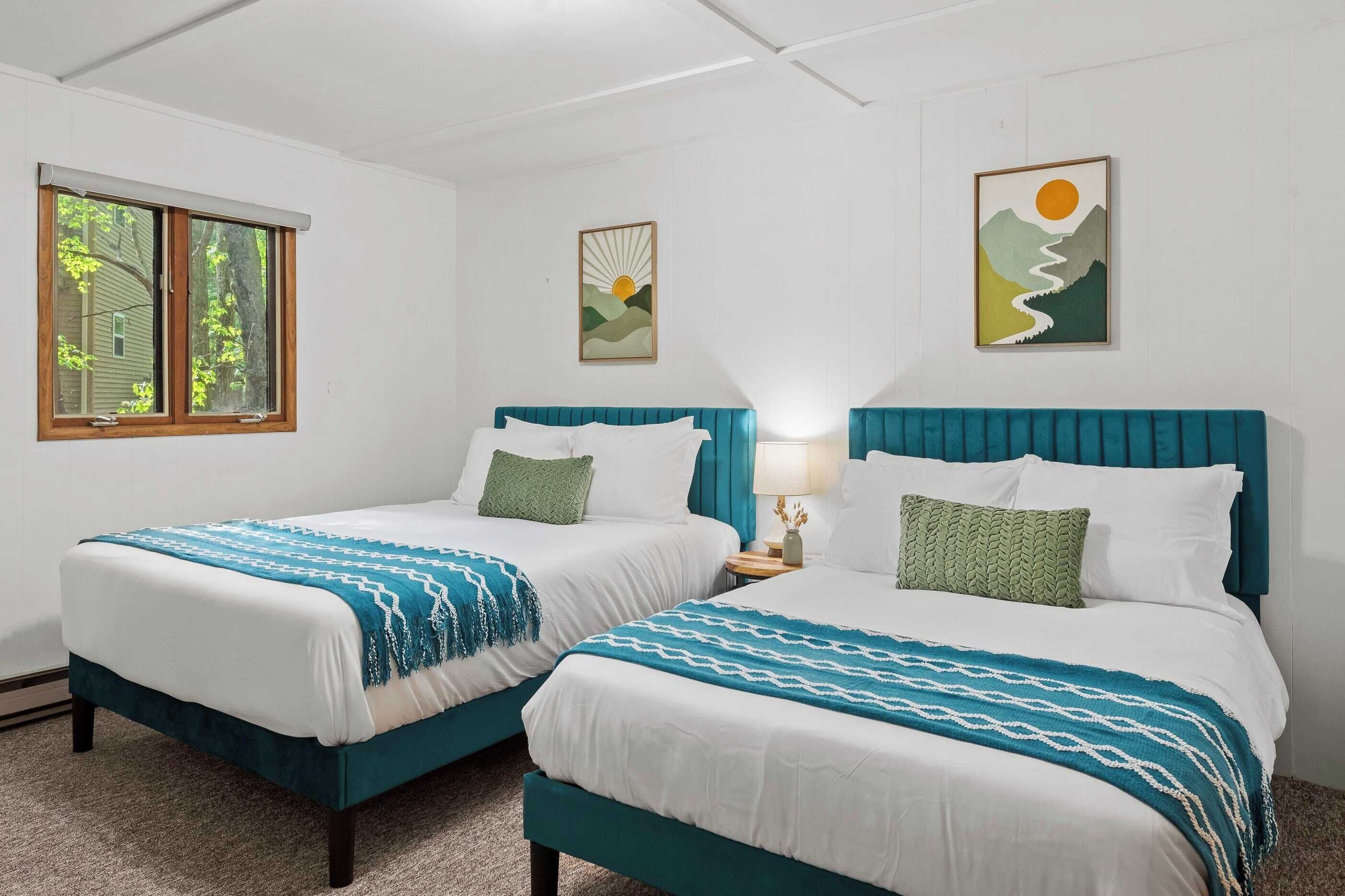 Quarto de hotel com duas camas, cada uma com cabeceira azul e roupa de cama branca, uma janela com vista para áreas verdes e duas obras de arte penduradas na parede.
