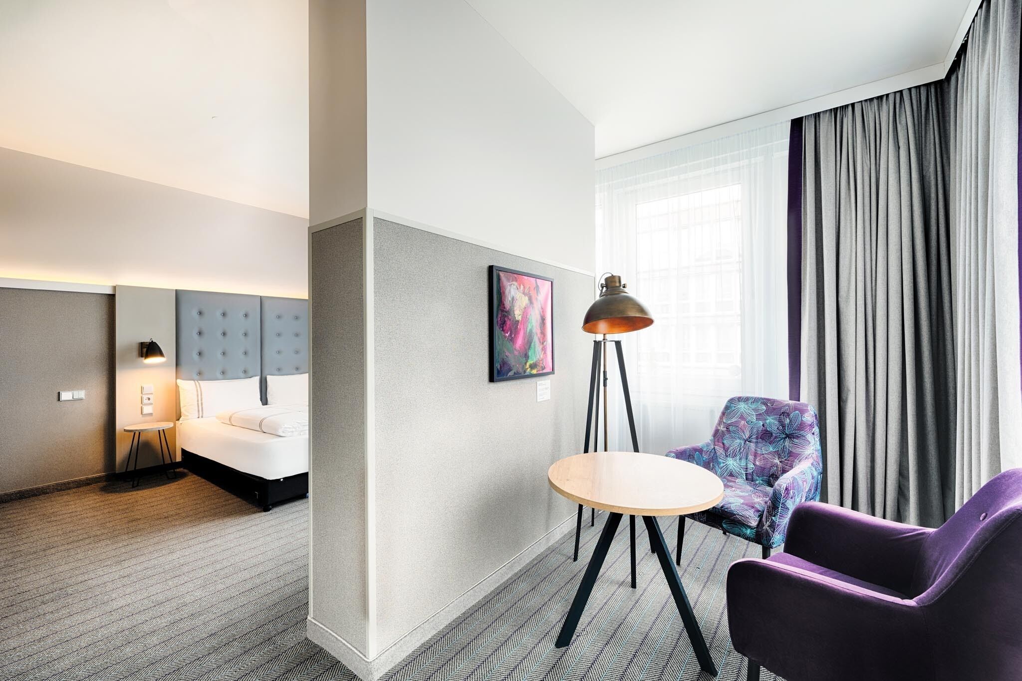 Quarto de hotel com cama, uma mesinha, duas cadeiras, um abajur e um quadro na parede.
