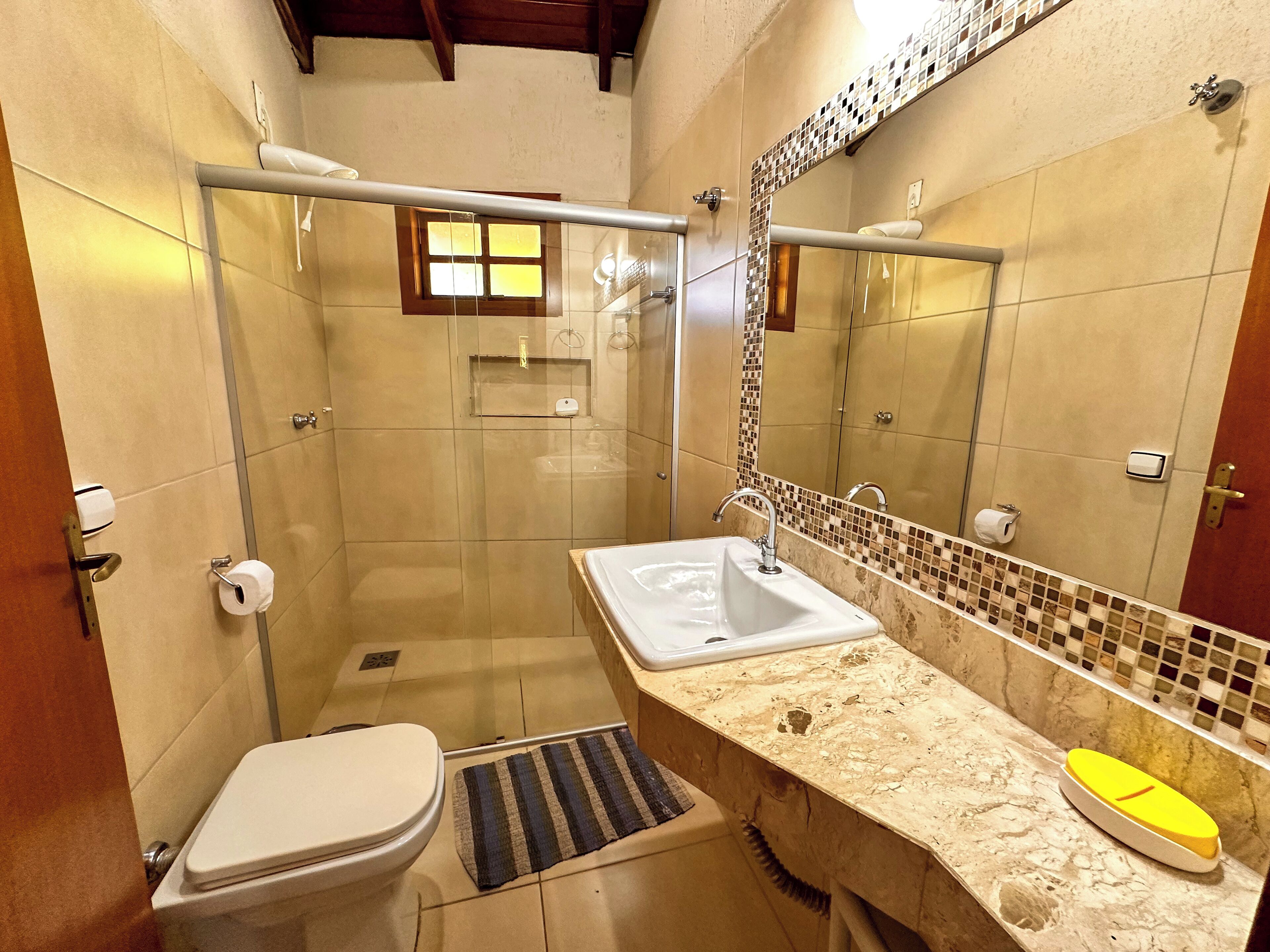Casa de banho com chuveiro, sanita, lavatório e espelho com painel de mosaicos.