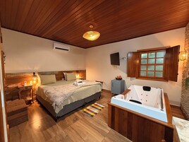 Quarto com cama, mesas de cabeceira, pequena escrivaninha, abajur, TV na parede, janela com persianas de madeira, banheira embutida e piso de madeira.