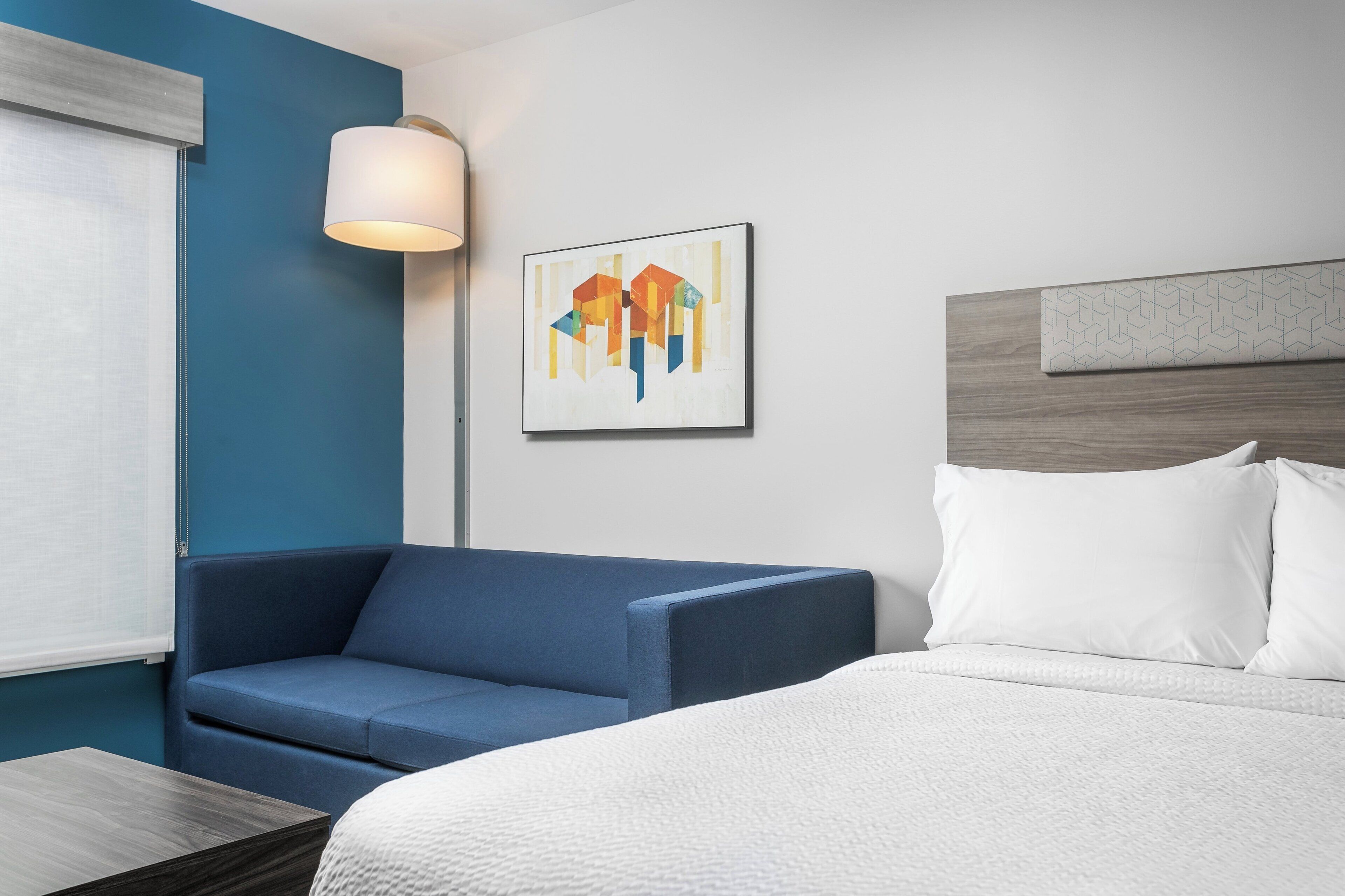 Quarto de hotel com uma cama, uma poltrona azul, uma luminária de parede e uma obra de arte geométrica.