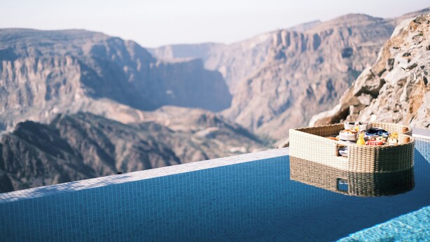 Piscina infinita com uma poltrona de vime e uma cesta de alimentos, com vista para uma paisagem montanhosa.