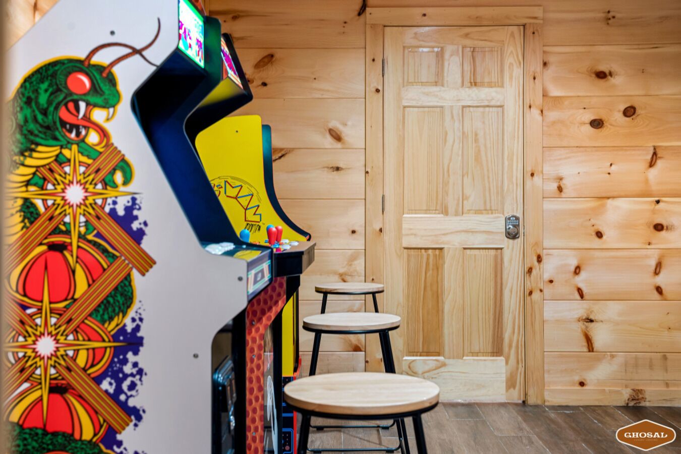 Interior de madeira com uma porta de madeira, uma máquina de jogos arcade colorida e duas mesas redondas.