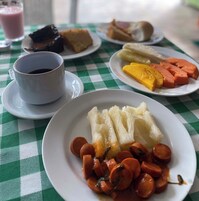 Uma mesa com toalha xadrez, uma chávena de café e pratos com comida, incluindo salsichas, batatas e uma fatia de bolo.