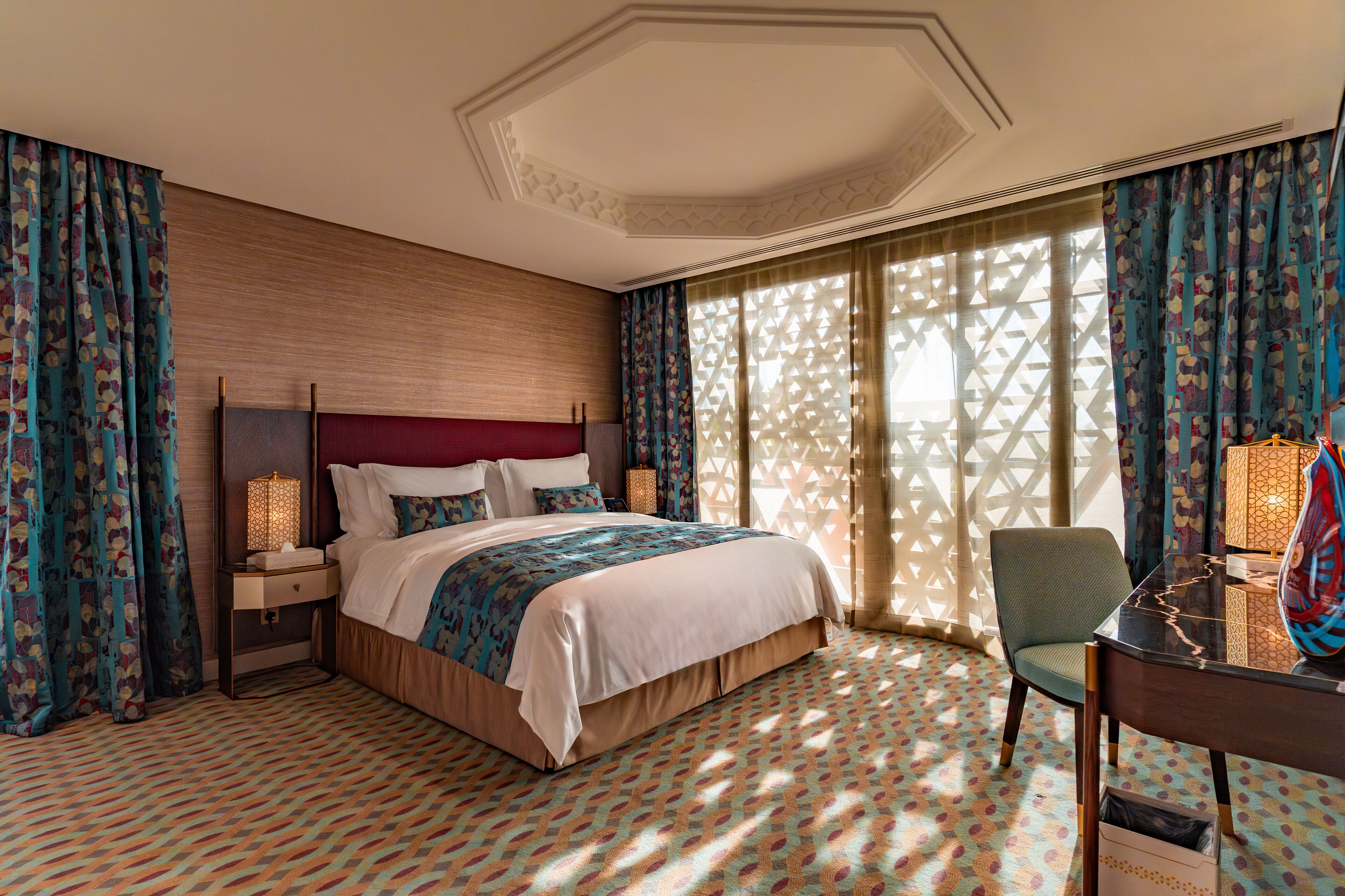 Quarto de hotel com uma cama grande, uma mesa de cabeceira com abajur, uma cadeira e uma escrivaninha com luminária.