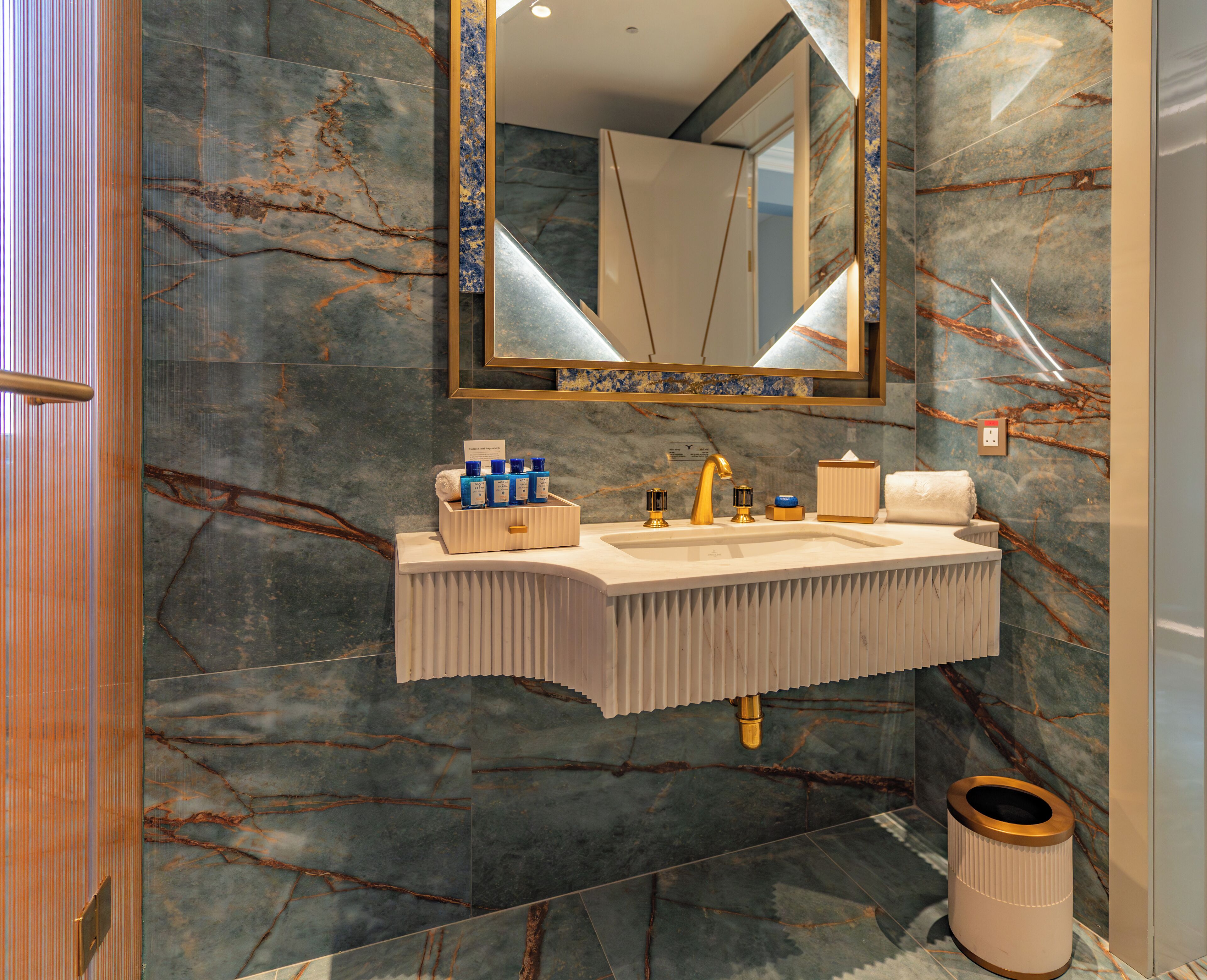 Casa de banho com parede de mármore, lavatório com produtos de higiene e um espelho.