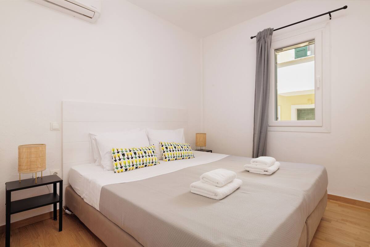 Cama bem arrumada com roupa de cama branca e cortina cinza, luminária de cabeceira e uma pequena mesa com abajur.