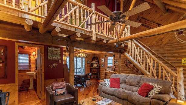 Interior de cabana com escada de madeira, sala de estar com sofá e poltrona, área de jantar e banheiro.