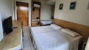 Quarto de hotel com cama, escrivaninha com televisão, guarda-roupa e banheiro pequeno.