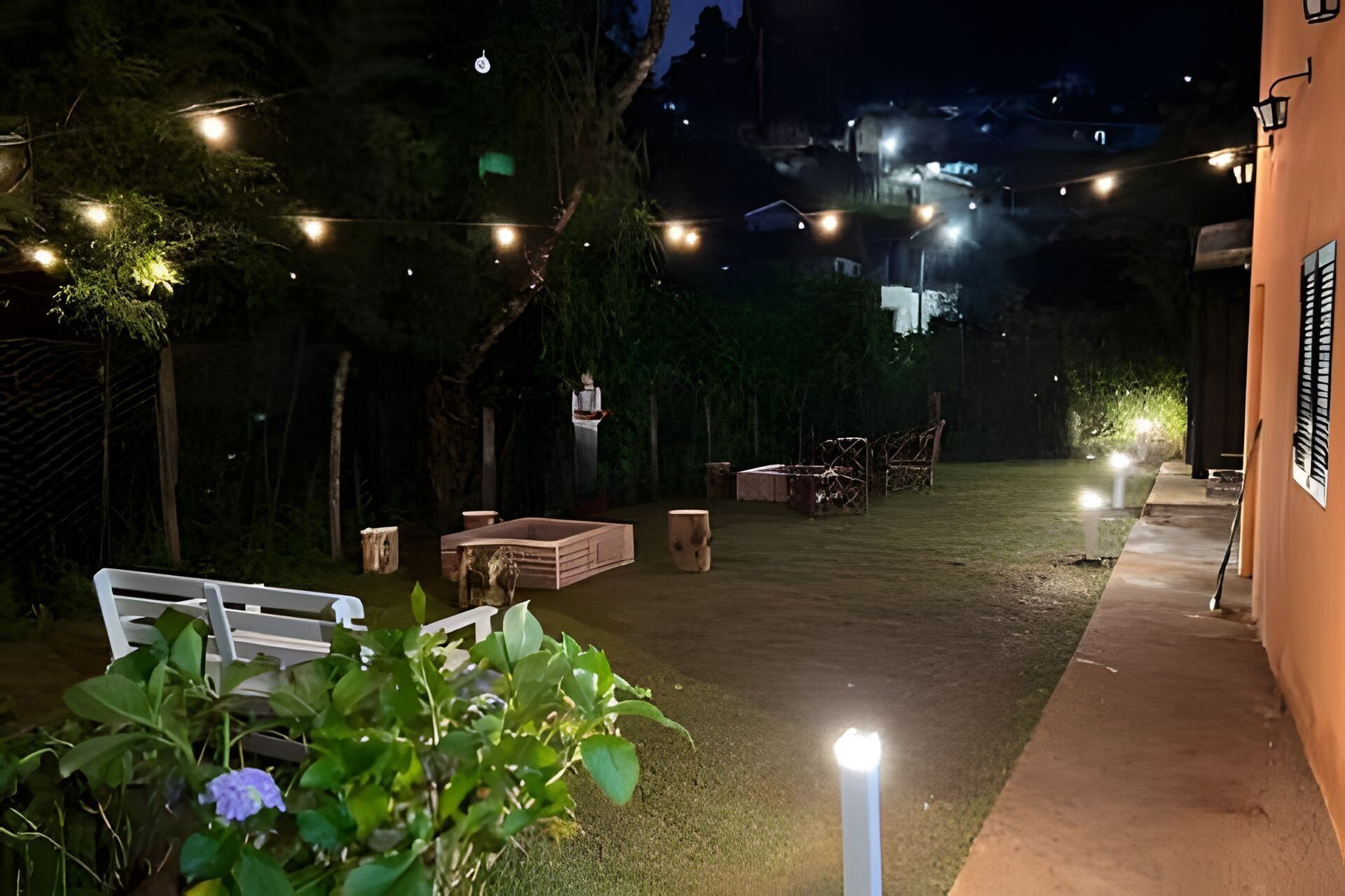 Uma área de pátio externa bem iluminada, com luzes pendentes, gramado e algumas estruturas de madeira.