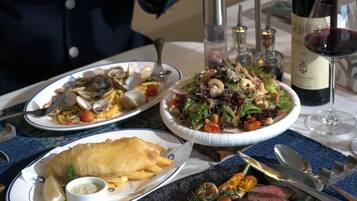 Uma mesa de jantar com diversos pratos, incluindo frutos do mar, salada e bife, juntamente com copos e garrafas de vinho.