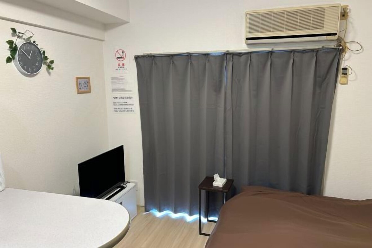 Quarto de hotel com uma cama, uma mesa com um laptop, um relógio e uma cortina.