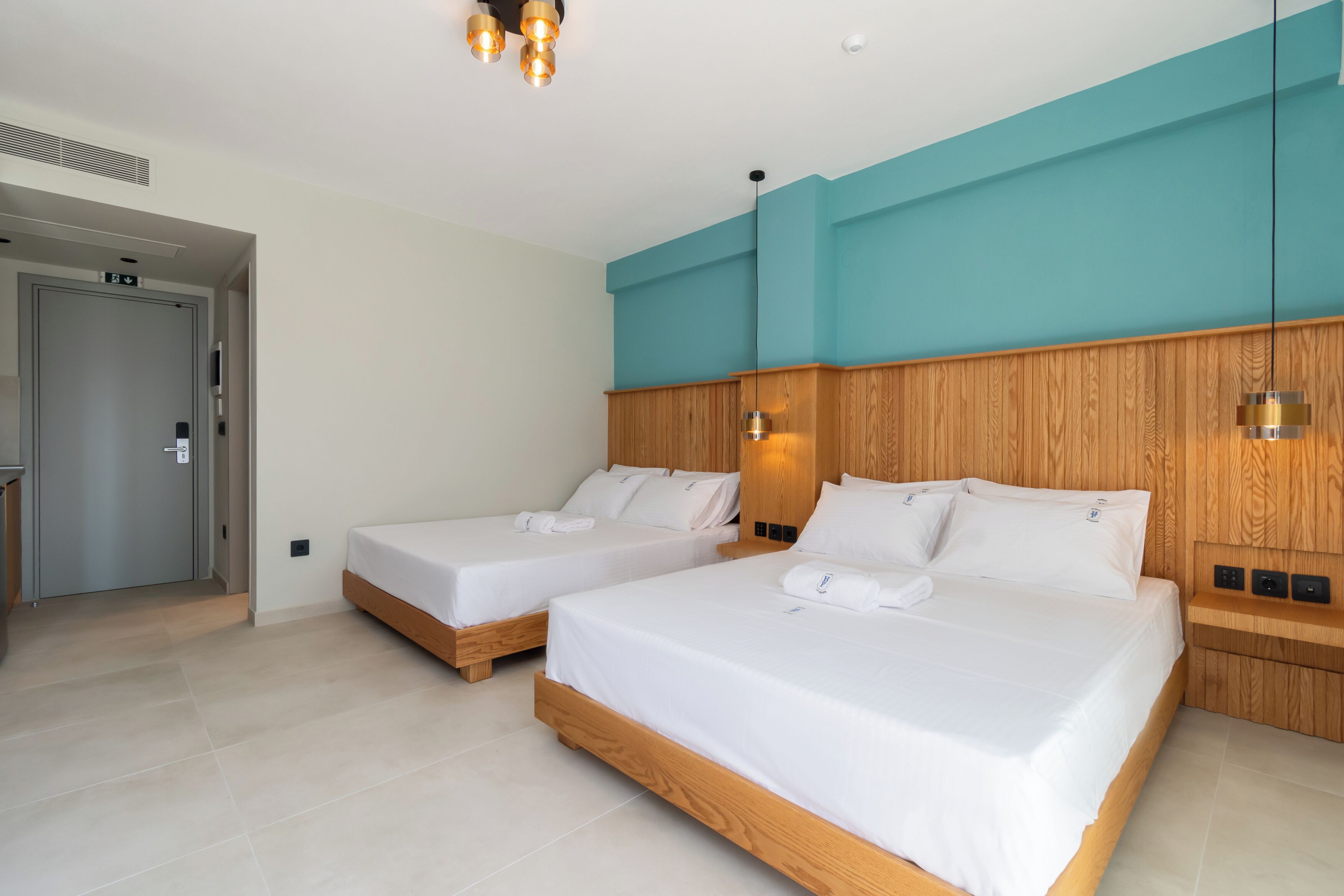 Quarto de hotel com duas camas, cabeceiras de madeira e uma parede de destaque em tons de verde-água.