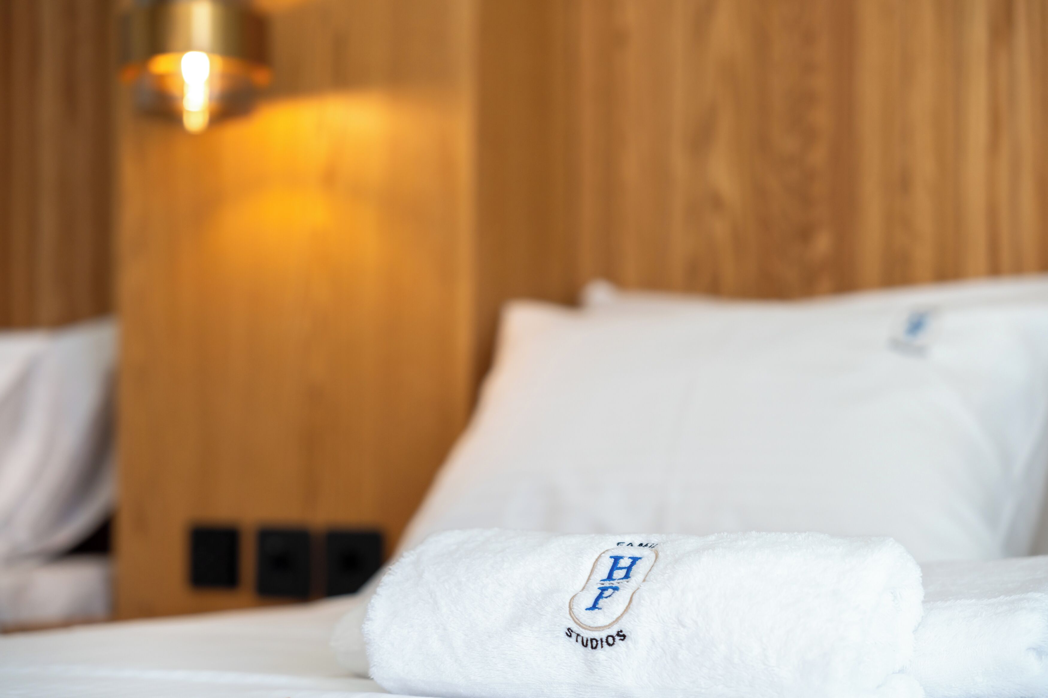 Cama bem arrumada com uma toalha branca com um logotipo, parede de madeira e luminária fixada na parede.