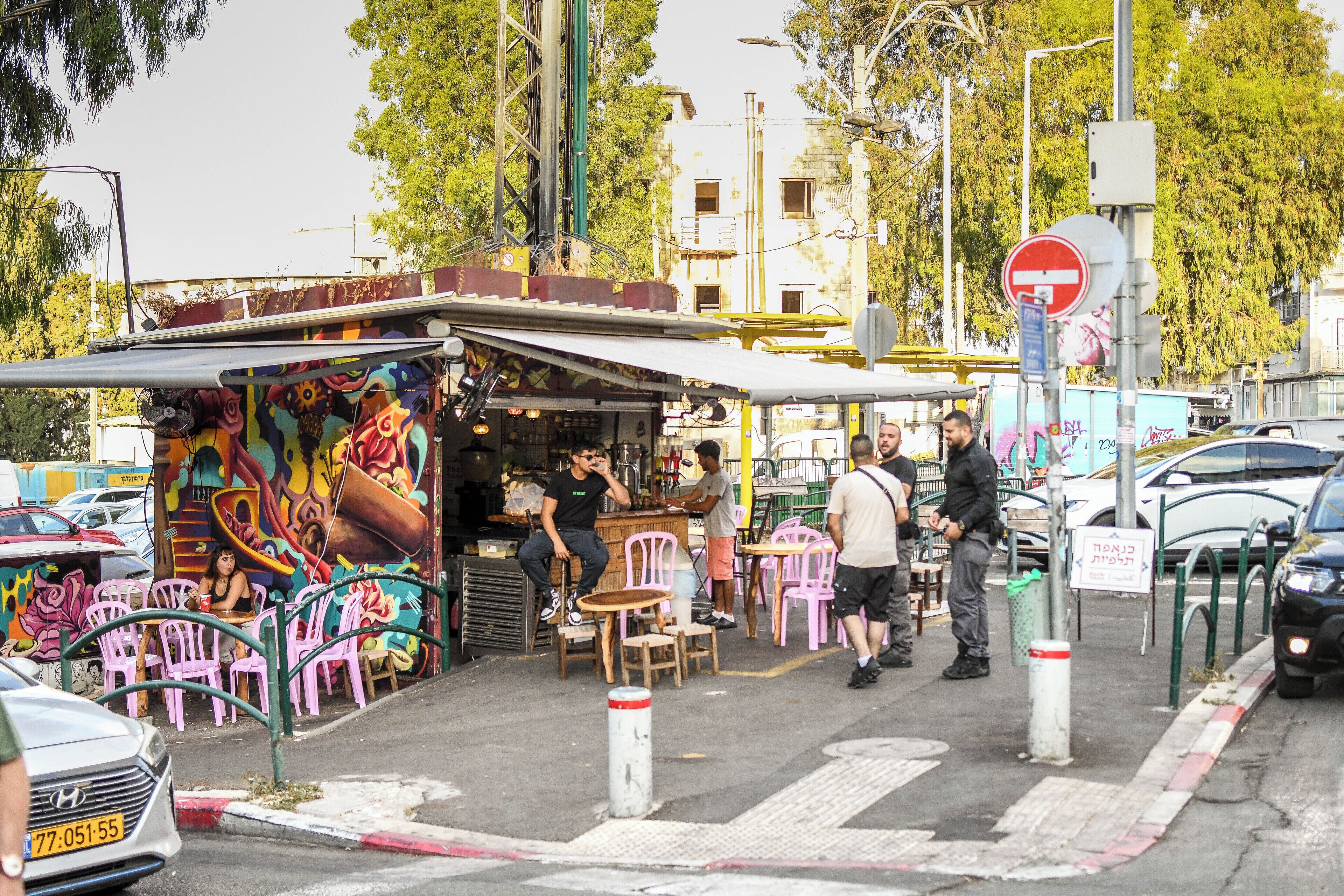 Um café à beira da rua com mesas ao ar livre, um mural e um sinal de proibido estacionar.