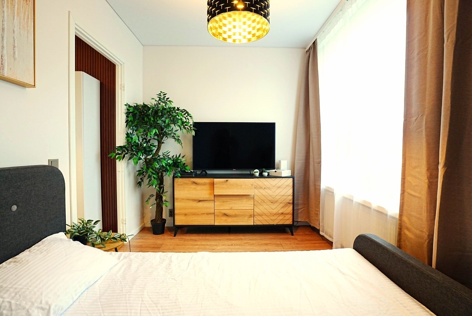 Quarto com cama, televisão sobre um armário de madeira, planta em vaso e janela com cortinas.