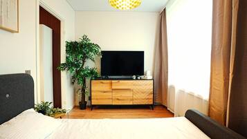 Quarto com cama, televisão sobre um armário de madeira, planta em vaso e janela com cortinas.