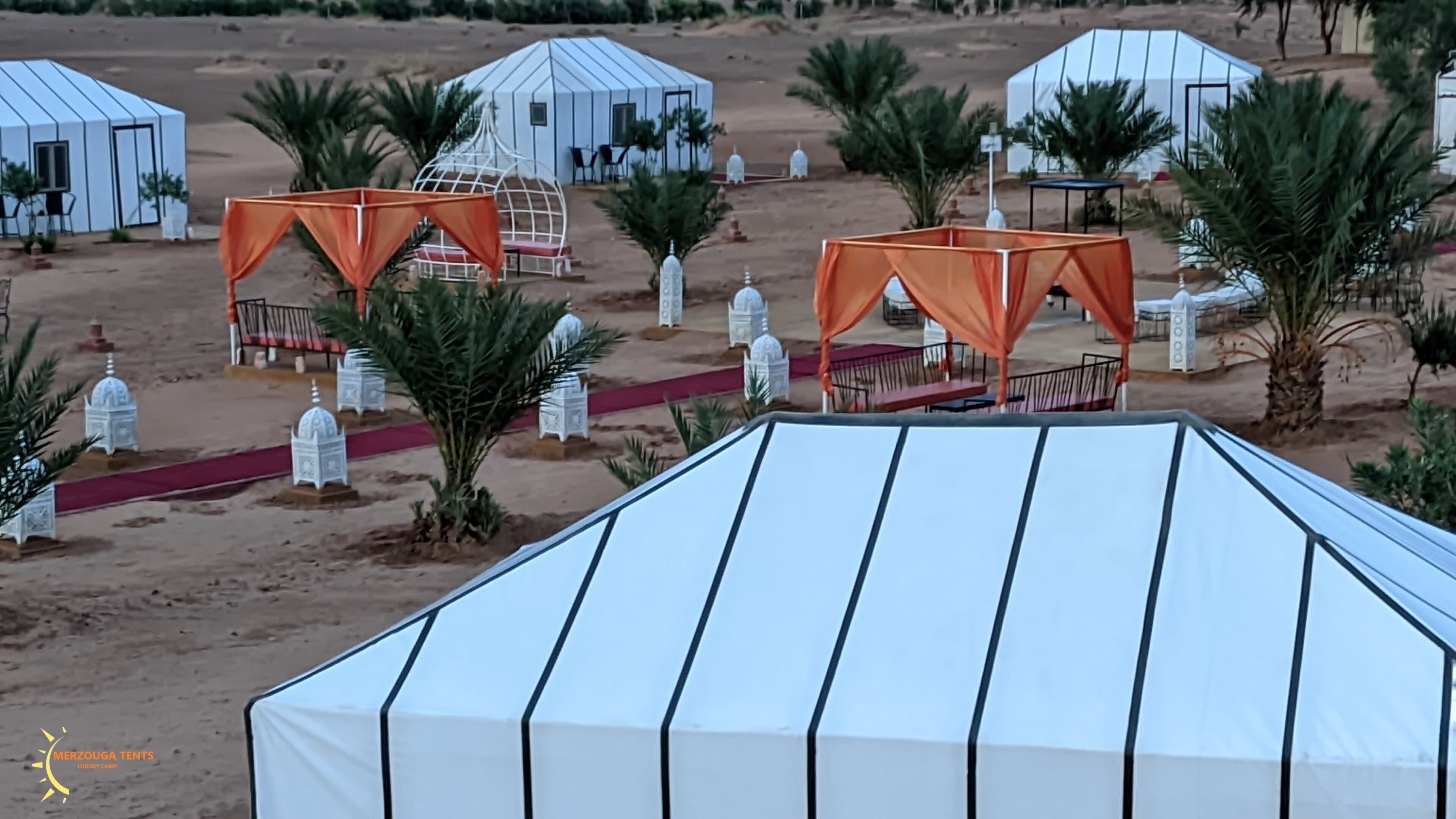 Acampamento no deserto com tendas brancas e toldos laranja, rodeado por palmeiras e lanternas brancas.