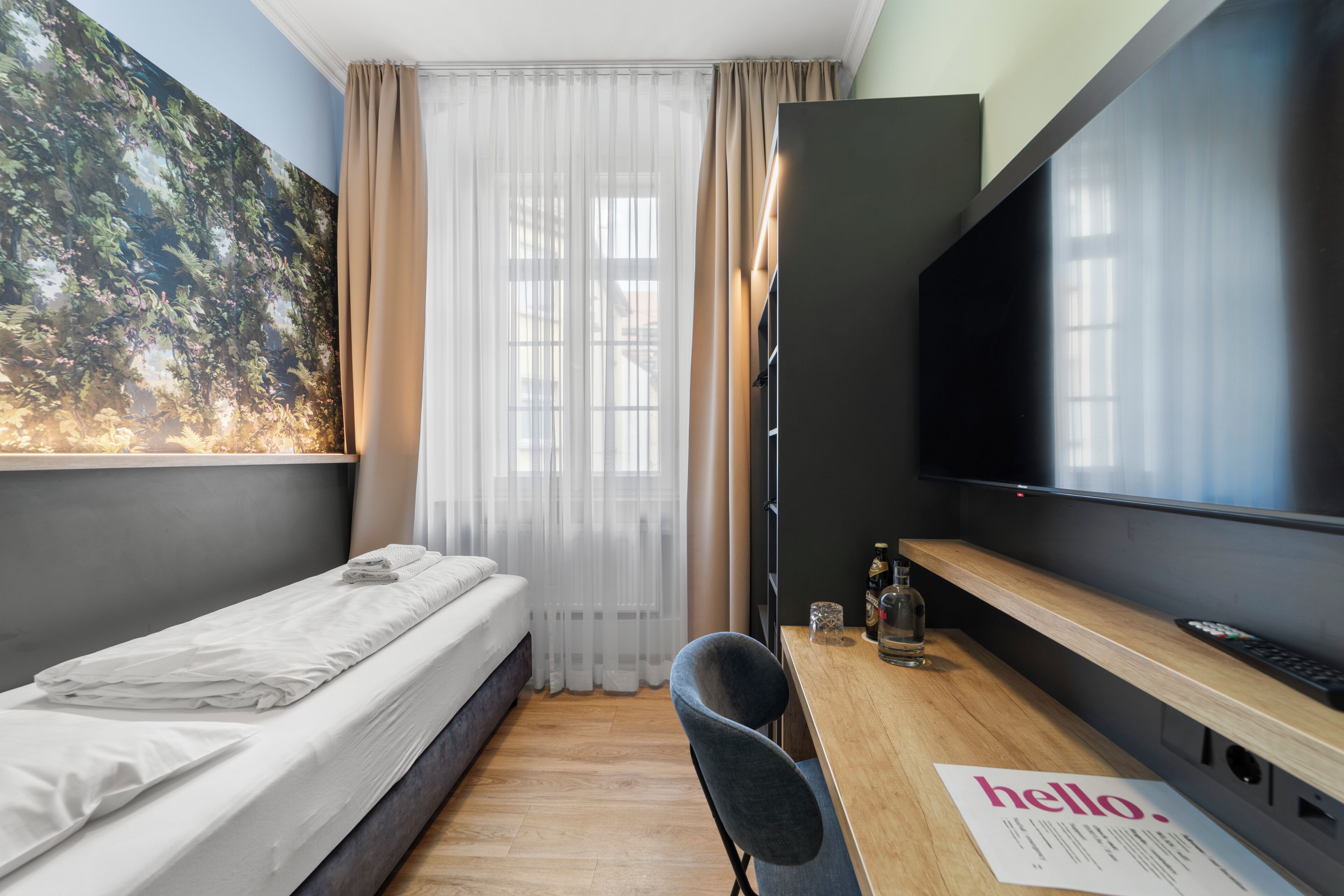 Quarto de hotel com cama, escrivaninha com cadeira, televisão e uma grande janela com cortinas.