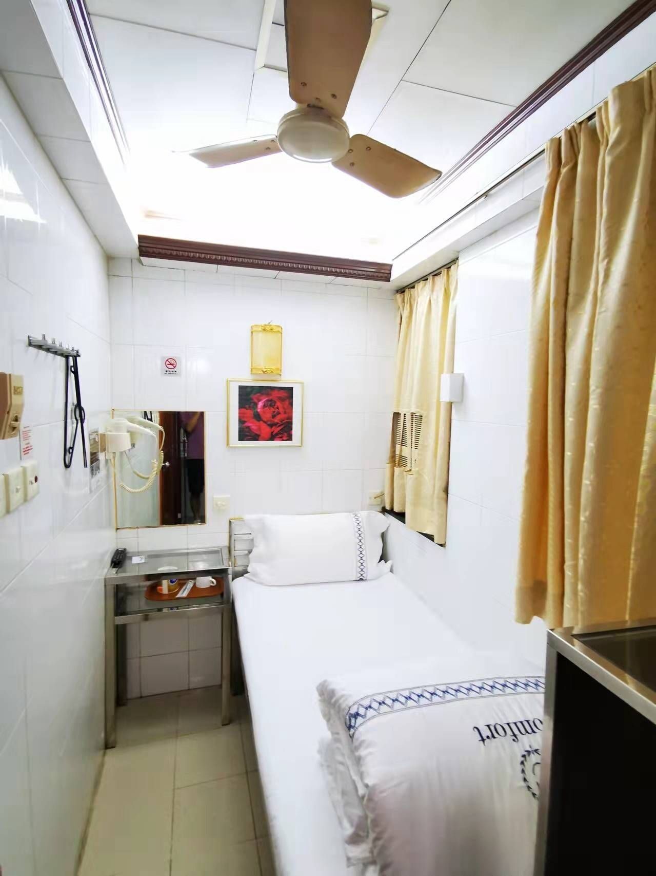 Quarto de hotel compacto com cama, escrivaninha, espelho, ventilador de teto e janela com cortinas.