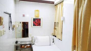 Quarto de hotel compacto com cama, escrivaninha, espelho, ventilador de teto e janela com cortinas.