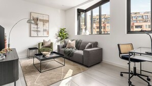 Uma sala de estar moderna com um sofá cinzento, uma mesa de centro, uma escrivaninha com cadeira, uma janela grande e um quadro na parede.