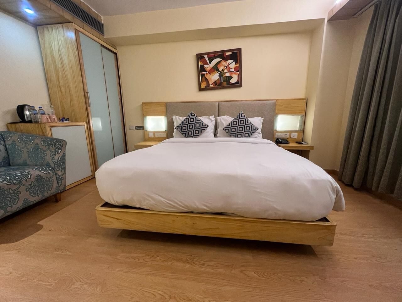 Quarto de hotel com uma cama grande, cabeceira de madeira, mesa de cabeceira com abajur e um quadro na parede.