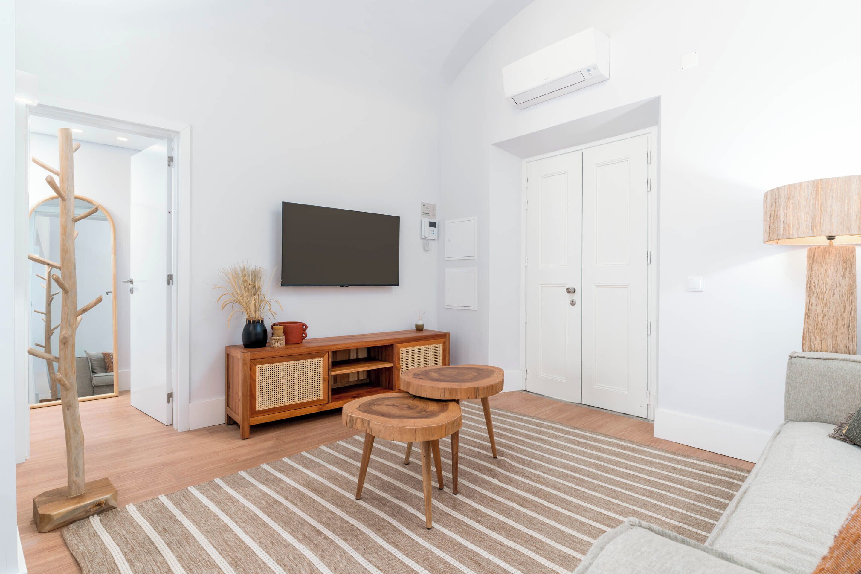 Uma sala de estar moderna com uma TV de tela plana na parede, uma mesa consola de madeira, duas mesas laterais redondas de madeira, um tapete listrado e um sofá.