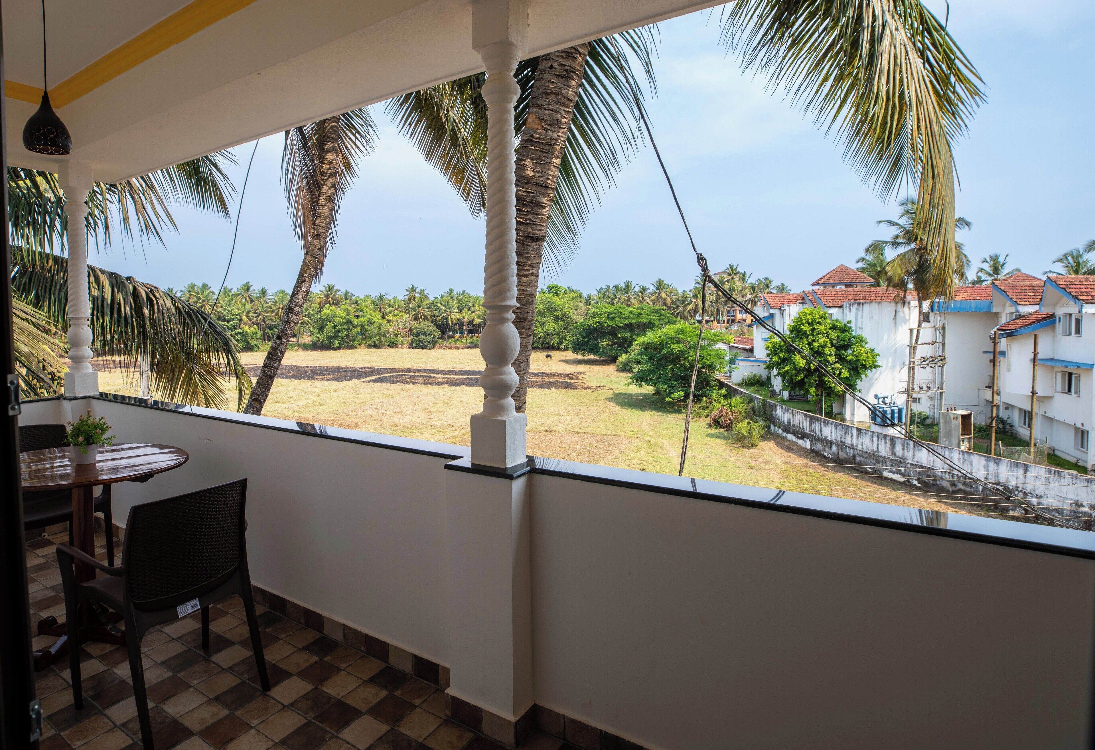 Uma varanda com piso xadrez, uma mesa e cadeiras. Há palmeiras e vista para uma área residencial com casas e uma praia.