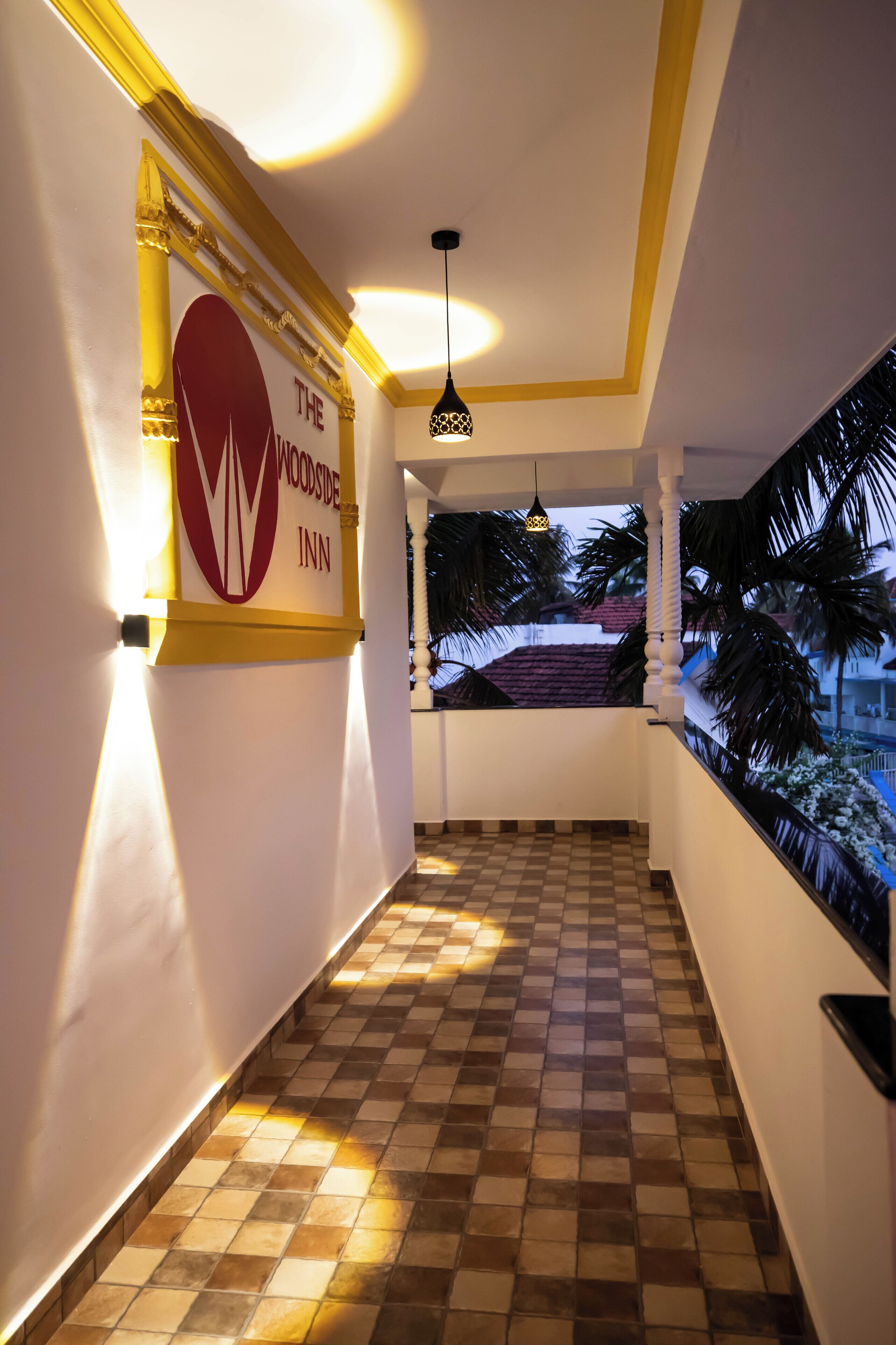 Um corredor de hotel com piso xadrez, uma placa com 'The Woodside Inn' e vista para palmeiras e um edifício ao fundo.