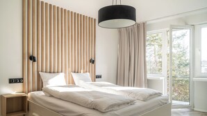 Quarto moderno com cama grande, ripas de madeira na parede, luminária pendente preta e vista para o exterior por uma janela ampla.