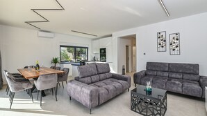 Uma sala moderna com um sofá cinzento, uma mesa de jantar de madeira e uma cozinha ao fundo.