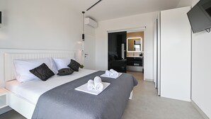 Quarto de hotel moderno com uma cama grande, televisão de tela plana e banheiro visível através de uma porta aberta.