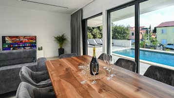 Uma sala de estar moderna com uma mesa de jantar de madeira, sofá cinza e uma grande tela de TV. Uma piscina é visível pelas grandes janelas.